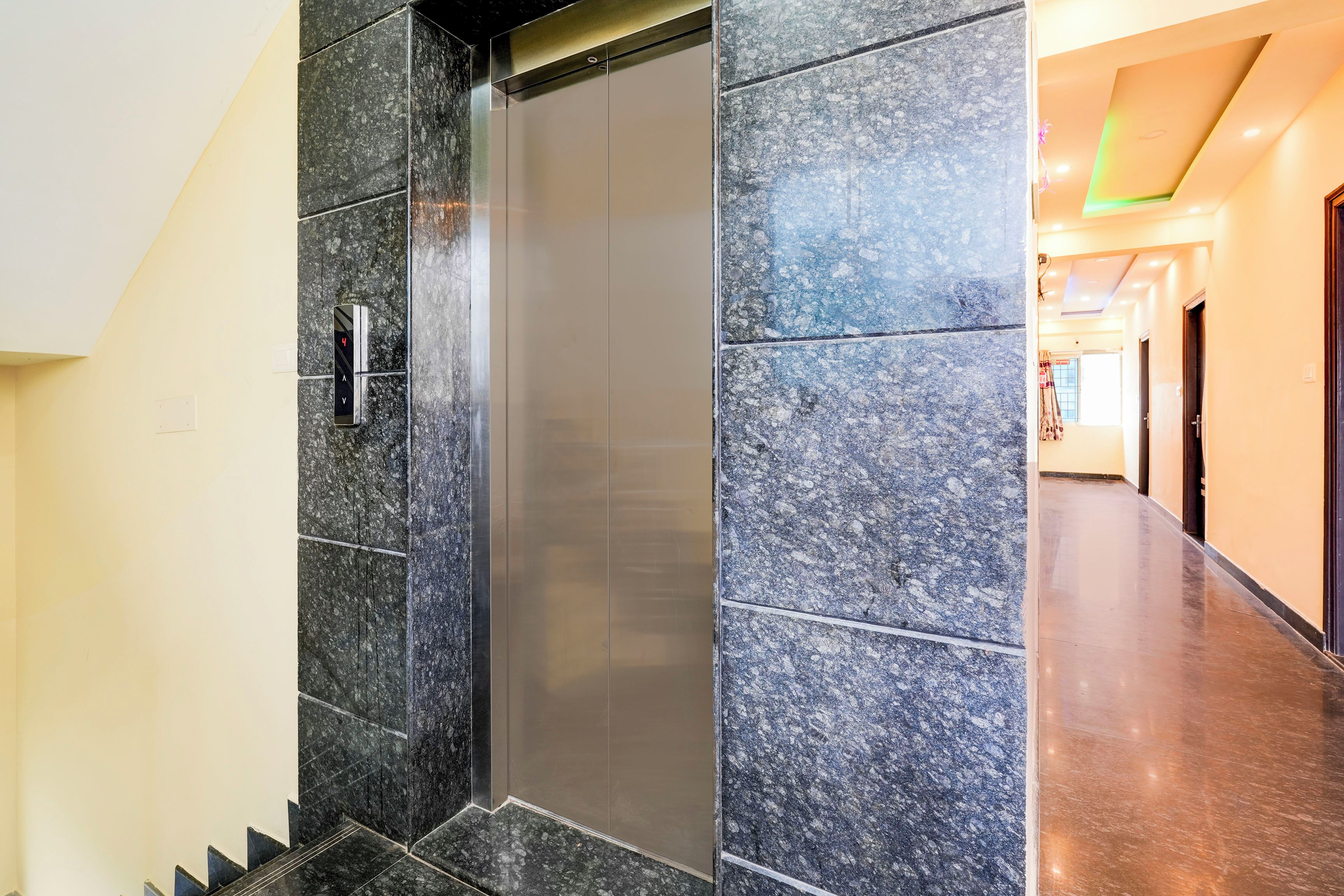 Um elevador moderno com acabamento metálico, ao lado de um corredor com piso lustroso e paredes com padrão pontilhado.