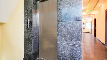 Um elevador moderno com acabamento metálico, ao lado de um corredor com piso lustroso e paredes com padrão pontilhado.