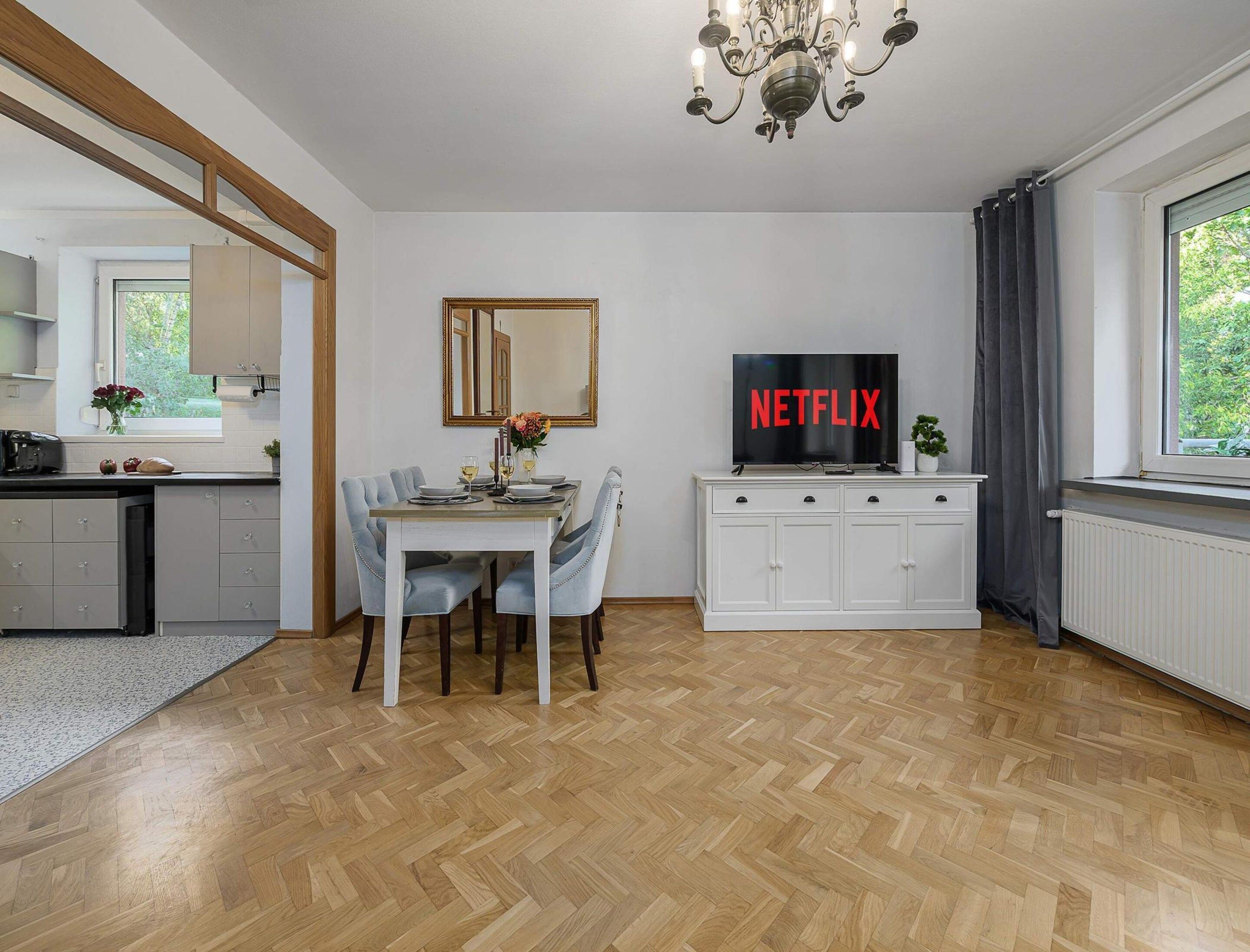 Uma sala de estar moderna com área de jantar, uma TV exibindo 'NETFLIX' e uma janela grande com cortinas.