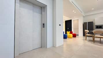 Interior moderno com elevador fechado, área de refeições com mesa e cadeiras, e poltronas de palha coloridas ao fundo.