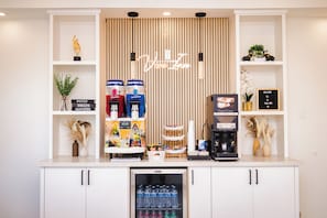 Uma área de cozinha compacta com cafeteira, dispensador de água e geladeira abastecida com bebidas.