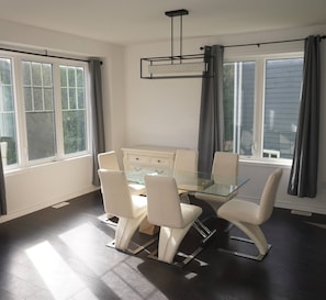 Uma área de refeições com mesa de vidro cercada por cadeiras, um armário branco e uma luminária pendente.