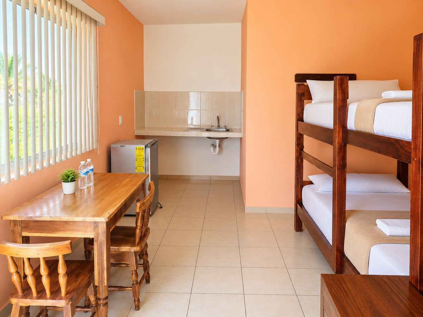 Quarto em dormitório com beliches, escrivaninha de madeira, cadeiras, pequeno espaço de cozinha e janela com persianas.