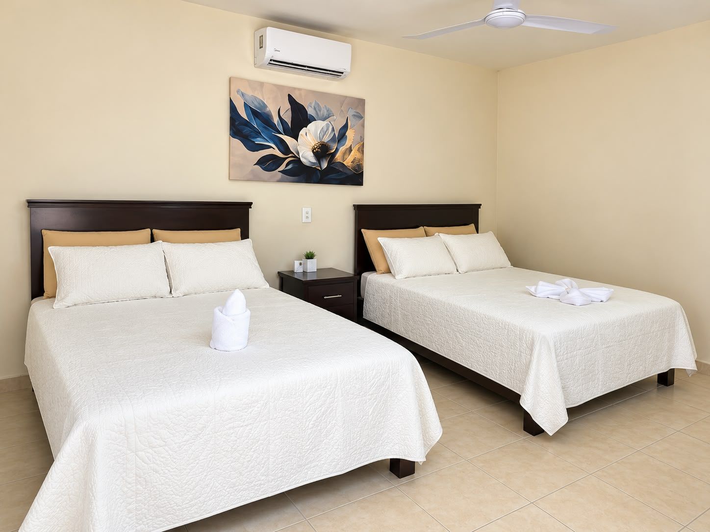 Quarto de hotel com duas camas, paredes bege, ventoinha no teto e um quadro.