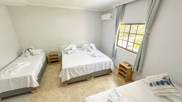 Quarto de hotel com duas camas de solteiro, uma janela grande com cortinas, uma pequena mesa de cabeceira de madeira e um aparelho de ar condicionado na parede.
