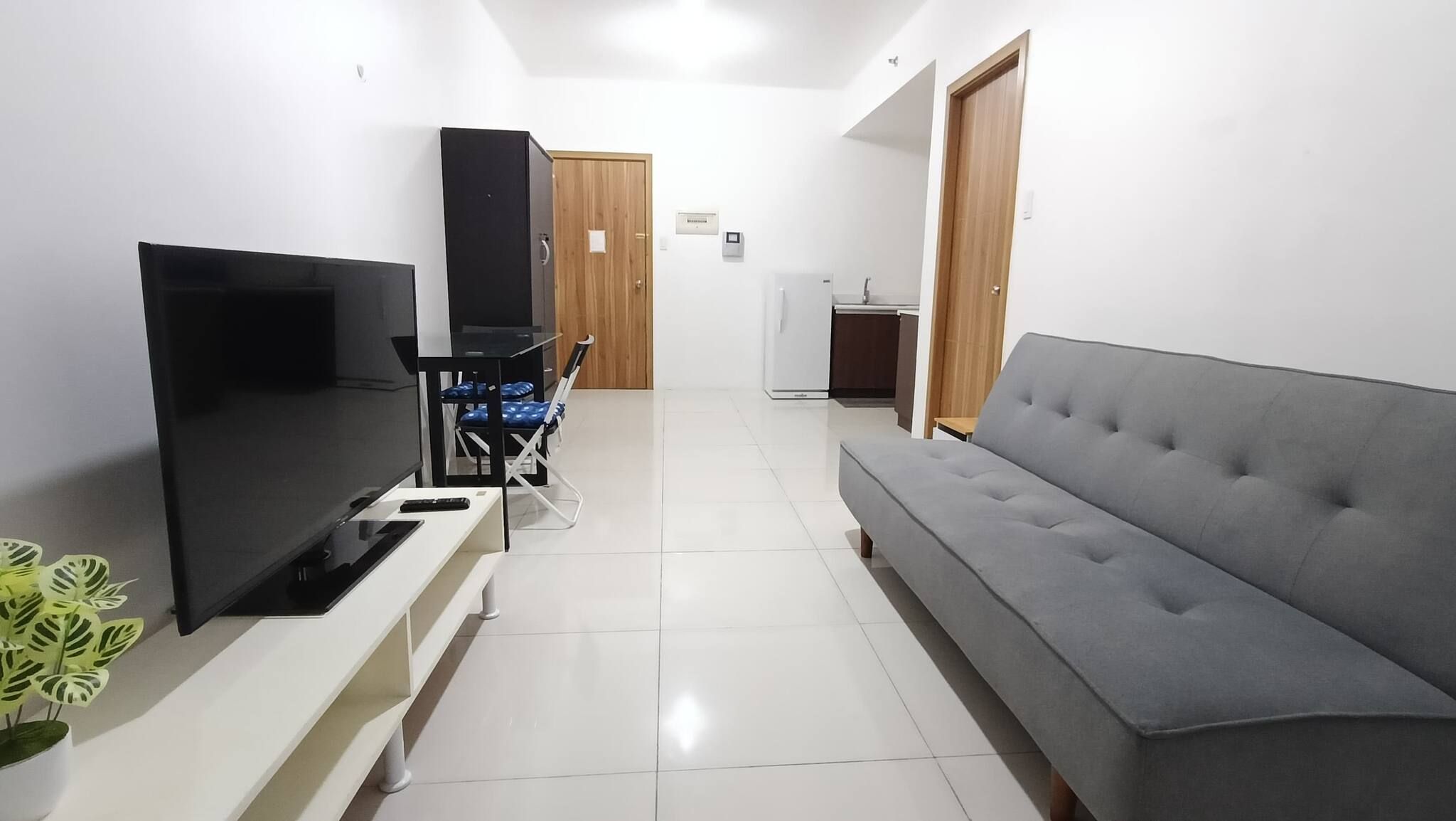 Sala de estar com sofá, televisão, mesinha e uma cadeira. Há uma porta e um termostato fixado na parede.