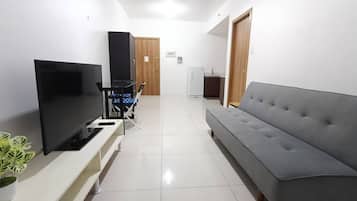 Sala de estar com sofá, televisão, mesinha e uma cadeira. Há uma porta e um termostato fixado na parede.