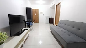 Sala de estar com sofá, televisão, mesinha e uma cadeira. Há uma porta e um termostato fixado na parede.