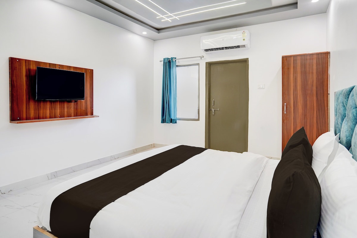 Quarto de hotel com cama, televisão de tela plana fixada em painel de madeira, ar condicionado, guarda-roupa e banheiro com espelho e cortinas azuis.