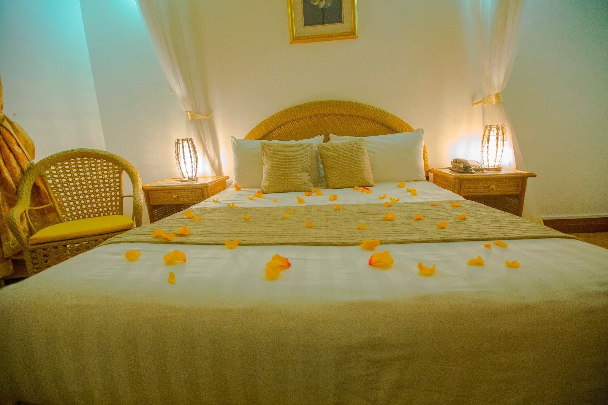 Cama bem arrumada com colcha e travesseiros brancos, uma cadeira de vime e uma mesa de cabeceira com abajur.