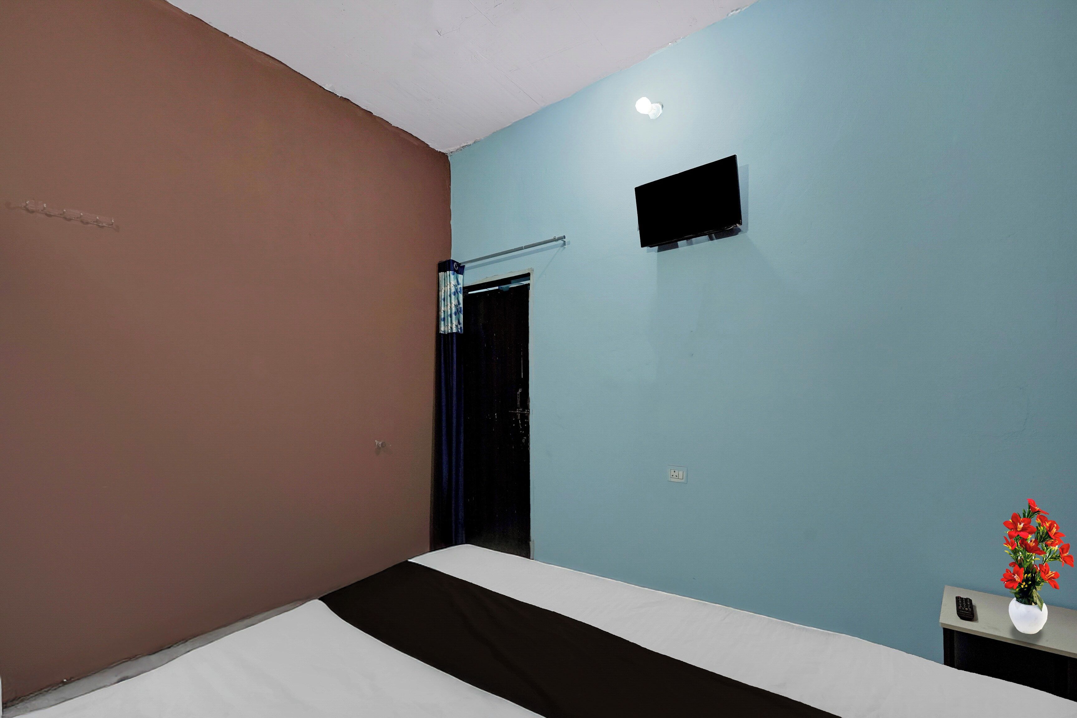 Um quarto com uma cama, uma mesinha com um vaso de flores, uma TV preta fixada na parede e uma porta.