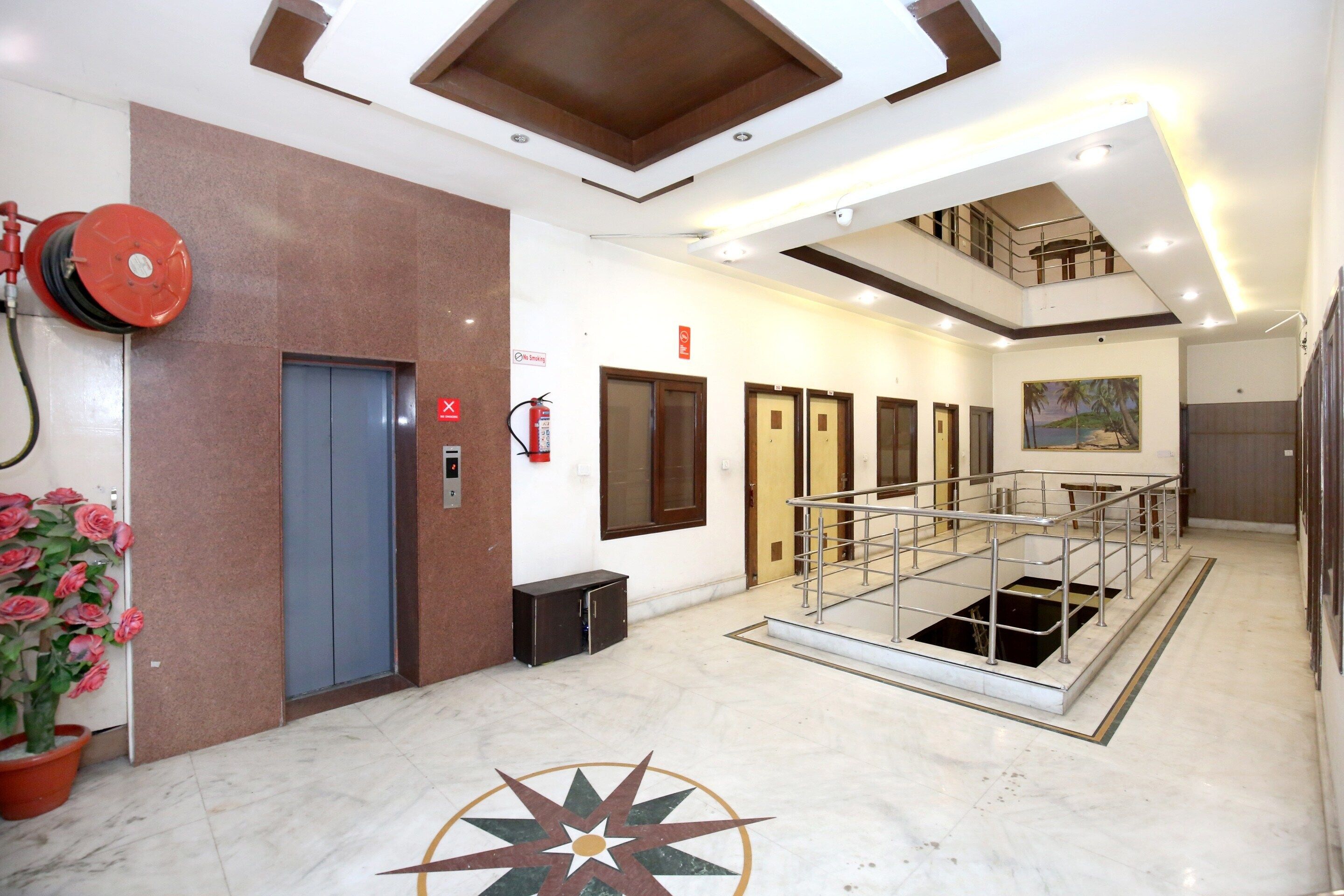 Um saguão de hotel com piso de mármore, um extintor de incêndio, uma placa de 'Proibido Fumar' e uma escadaria com corrimão de metal.