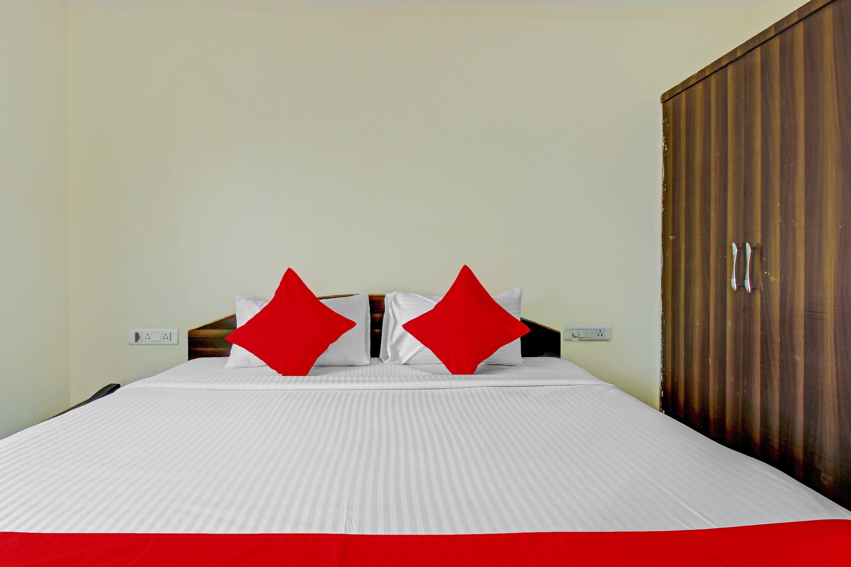 Uma cama com colcha branca e duas almofadas vermelhas, um guarda-roupa de madeira e uma mesa de cabeceira com abajur.