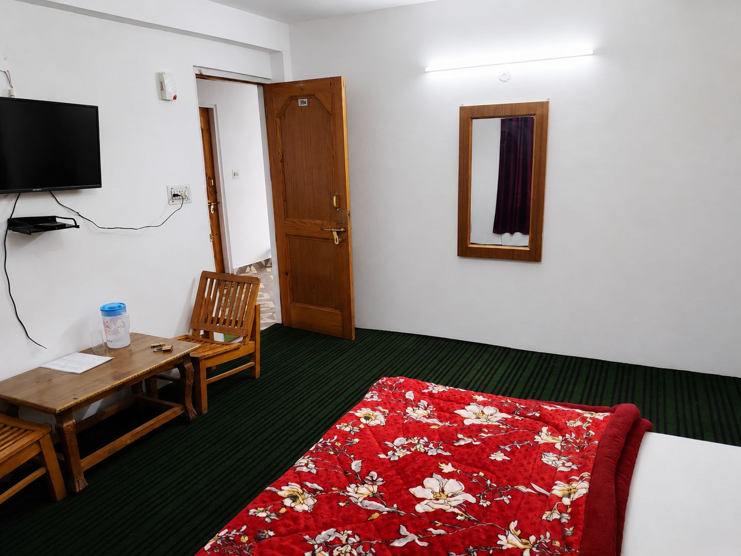 Quarto de hotel com cama, escrivaninha, cadeira, televisão e porta.