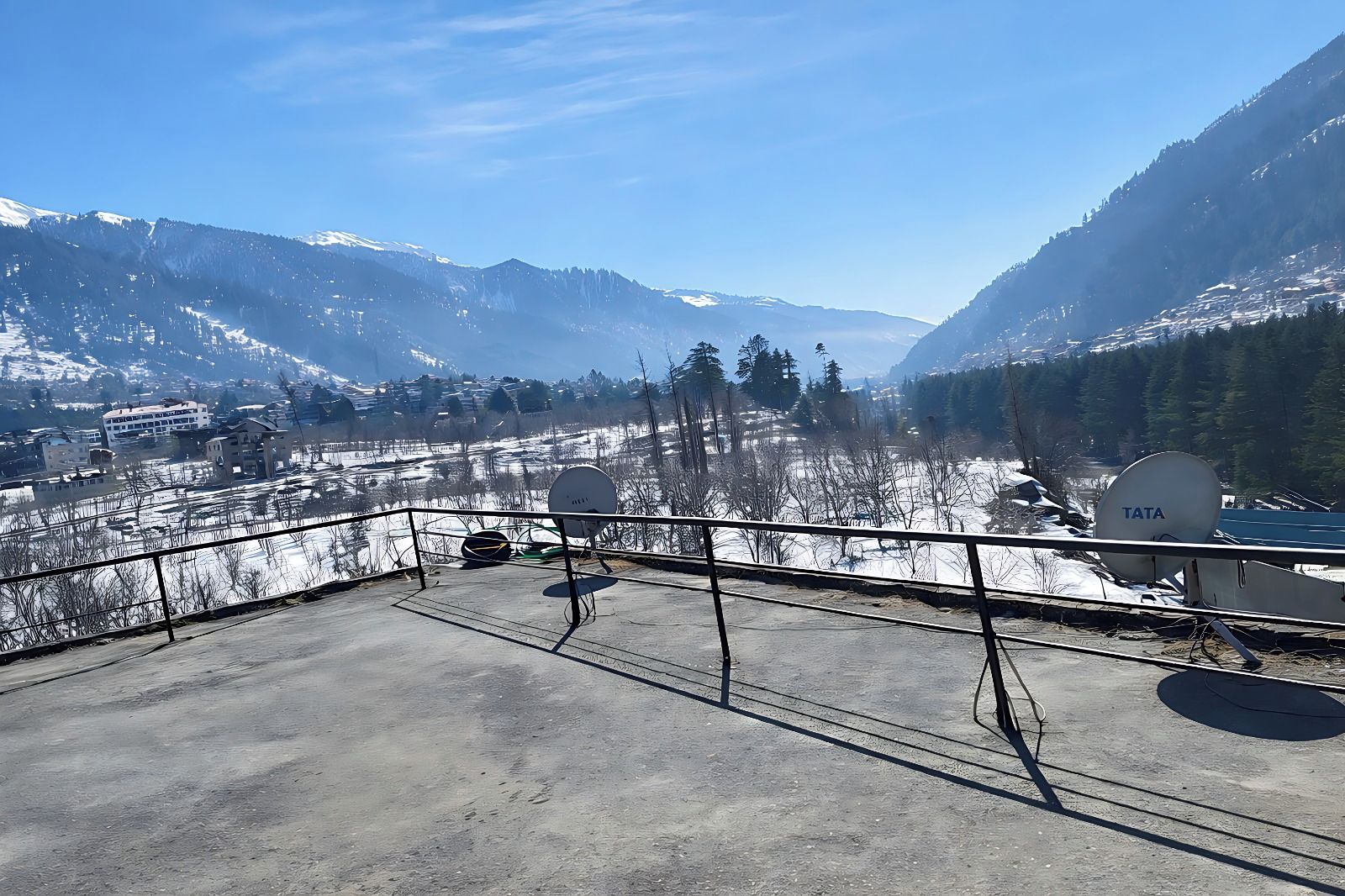 Uma plataforma de concreto com guarda-corpo de metal, com vista para um vale com montanhas cobertas de neve e alguns edifícios.