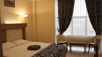 Quarto de hotel com cama, duas cadeiras, escrivaninha, luminária e janela com cortinas.