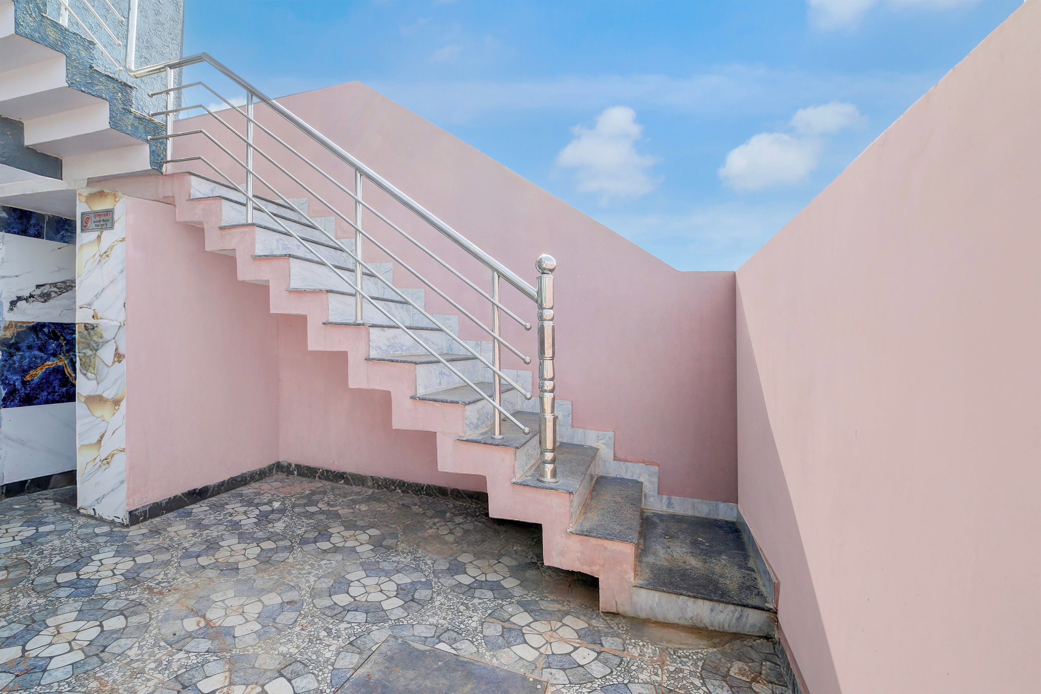 Escadaria com corrimão de metal que leva a um patamar, cercada por paredes cor-de-rosa e um piso de azulejos.