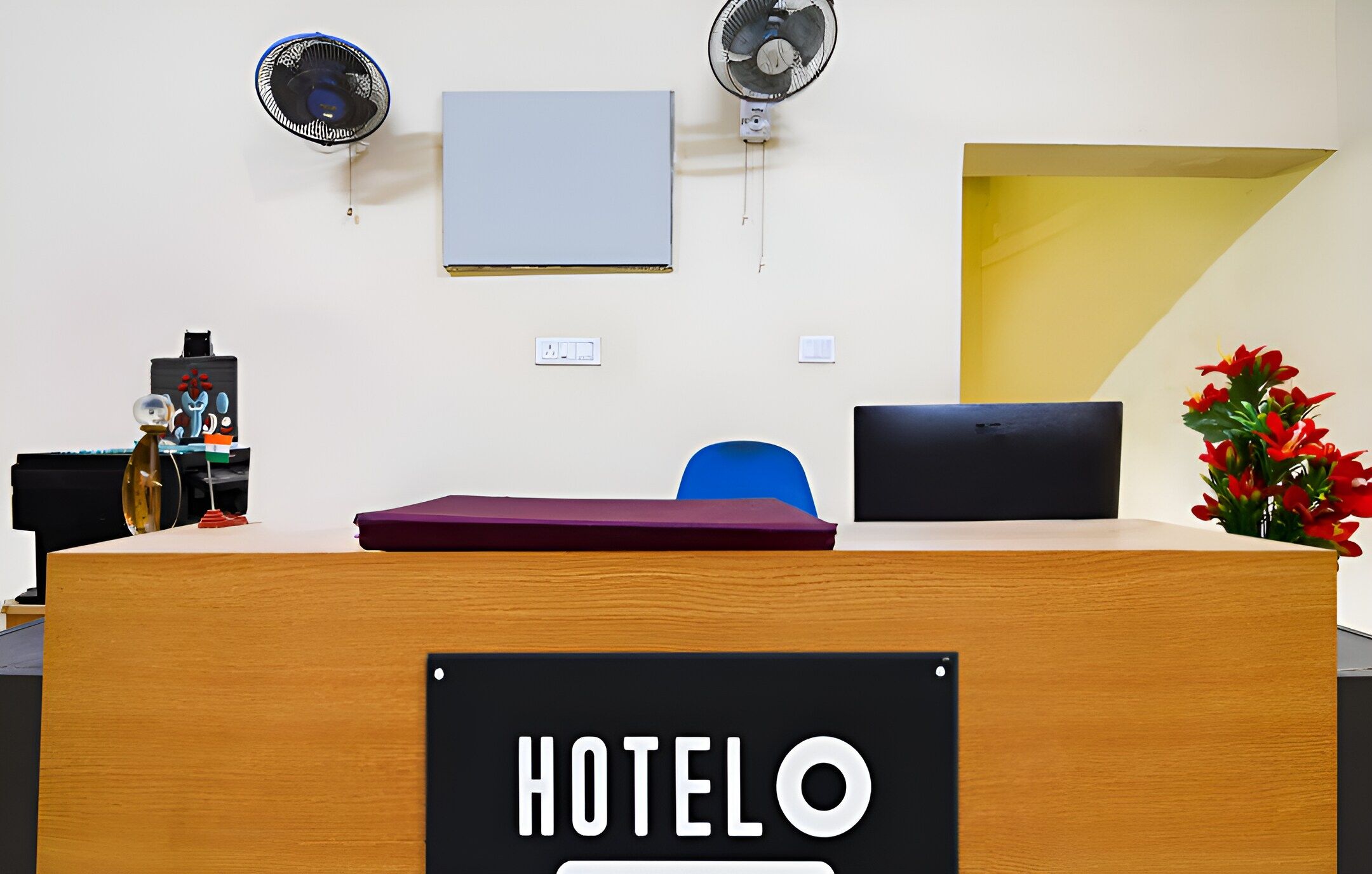 Uma receção de hotel com uma placa a indicar 'HOTELO', dois ventiladores fixados na parede, um computador portátil, um vaso com flores vermelhas e uma cadeira azul.