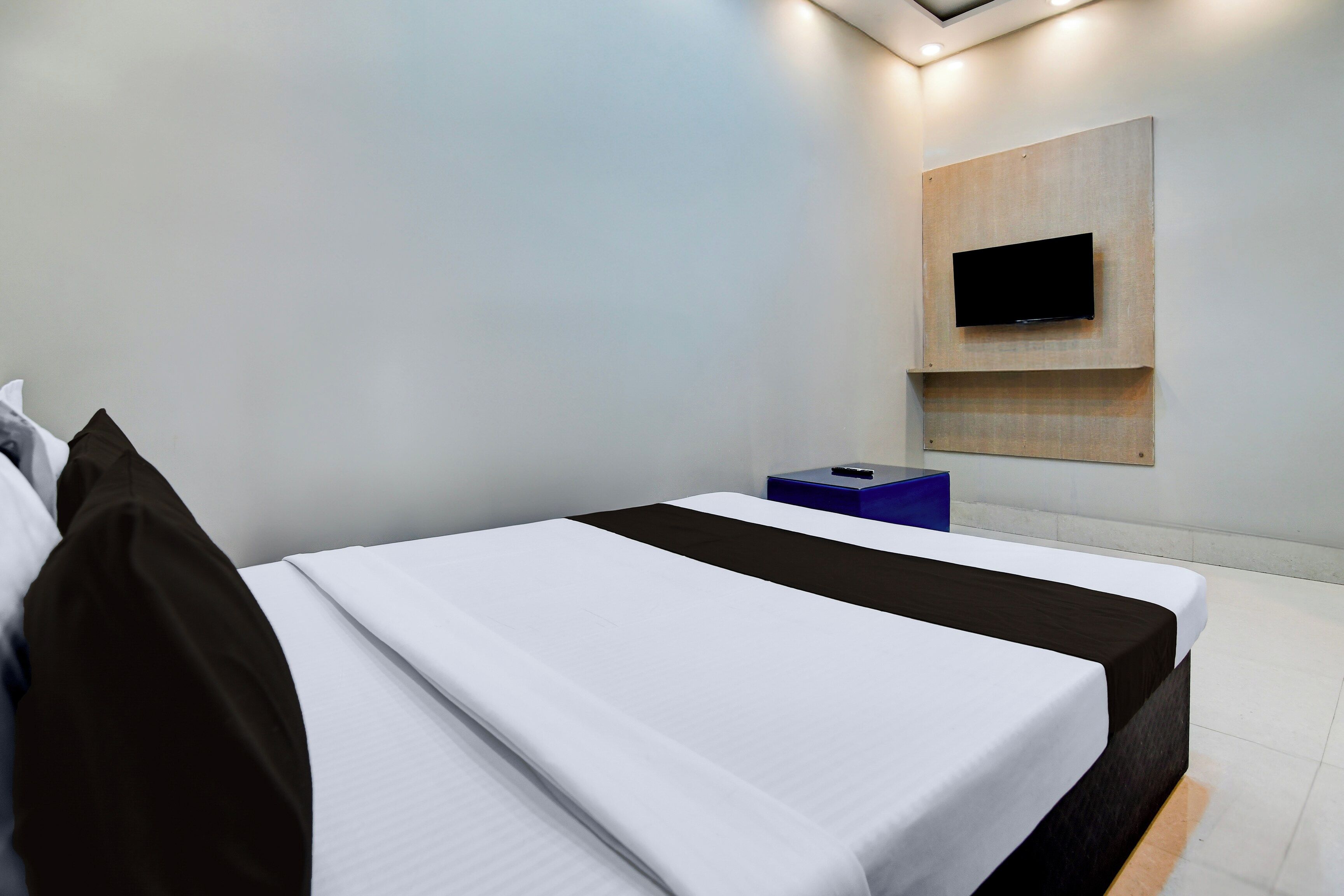Quarto de hotel moderno com uma cama, uma TV de tela plana na parede e um pequeno objeto azul na mesa de cabeceira.