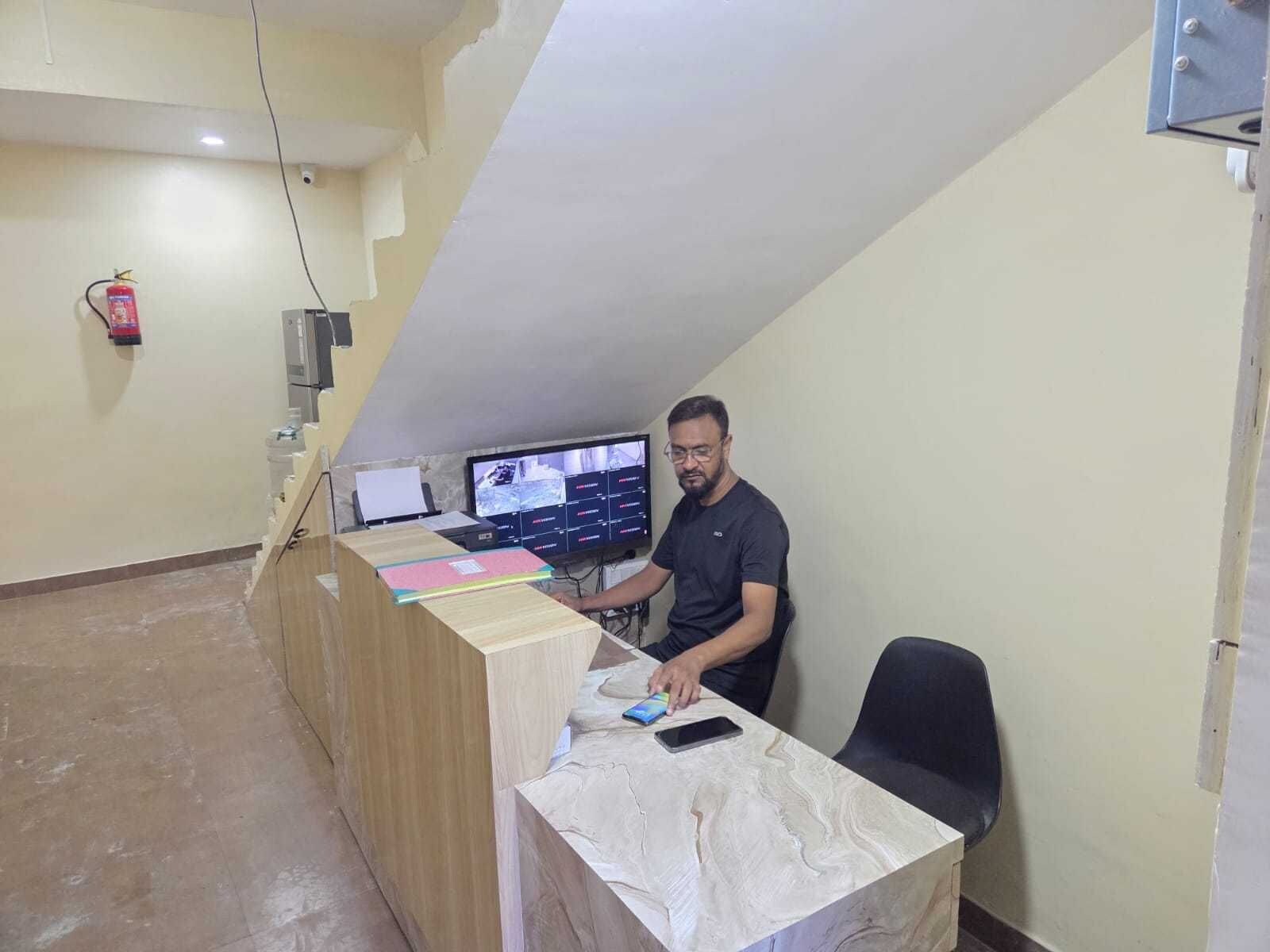 Um homem sentado em uma mesa com vários monitores de computador, um extintor de incêndio na parede e piso de azulejos.