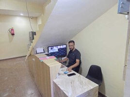 Um homem sentado em uma mesa com vários monitores de computador, um extintor de incêndio na parede e piso de azulejos.