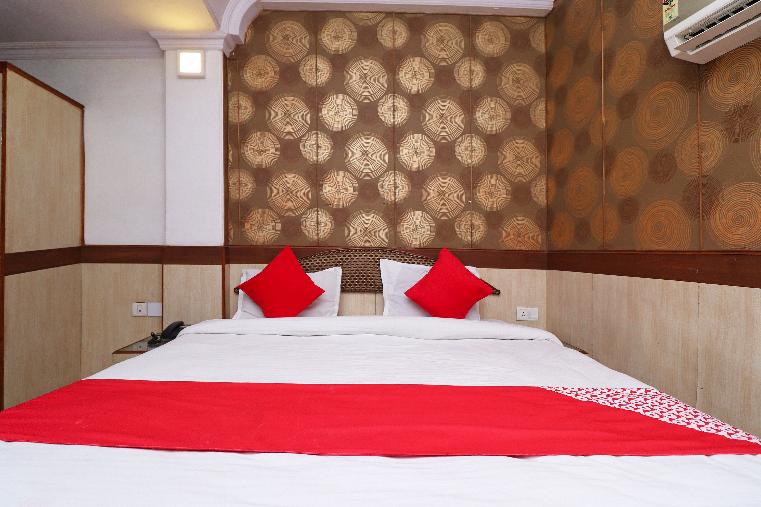 Quarto de hotel com uma cama grande, duas almofadas vermelhas, um edredom vermelho e branco, e um papel de parede estampado.