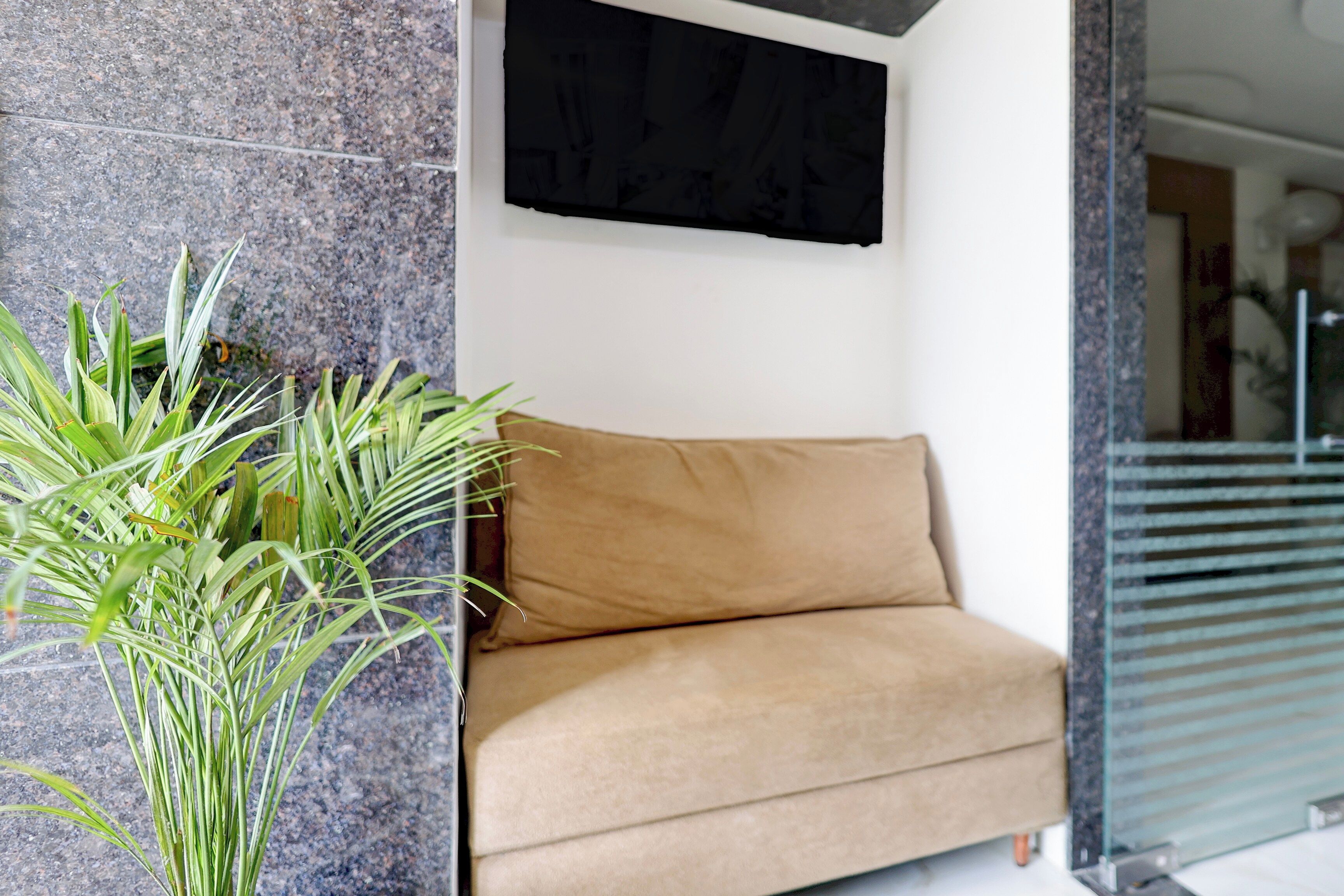 Quarto com sofá bege, planta em vaso e TV na parede.