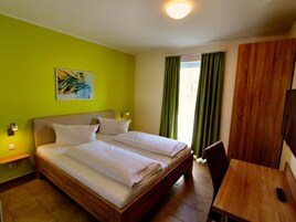 Quarto de hotel com cama de casal, escrivaninha, cadeira, quadro na parede e janela com cortinas.