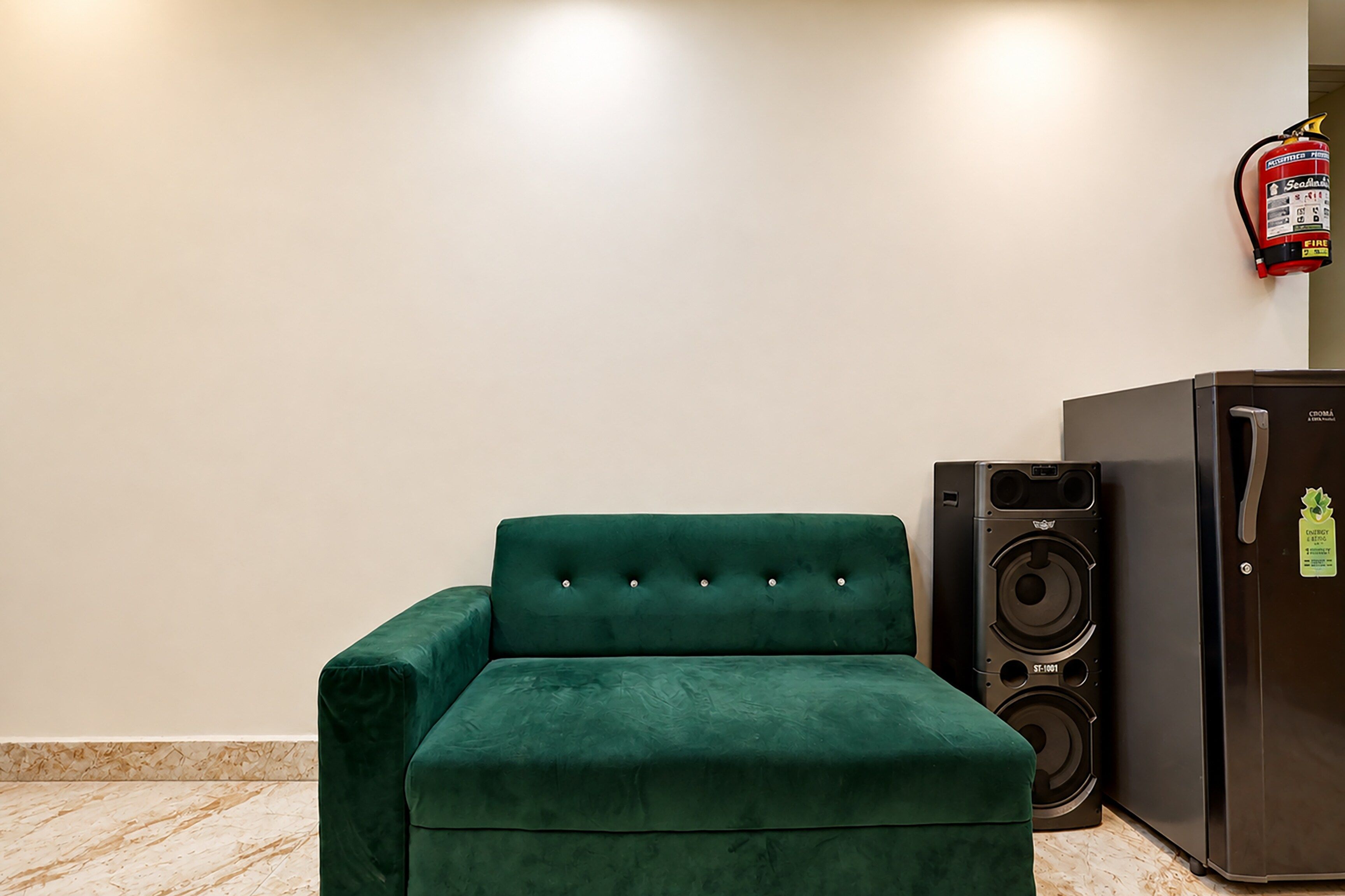 Uma cadeira estofada verde e um refrigerador preto em um quarto com paredes brancas e piso de mármore.