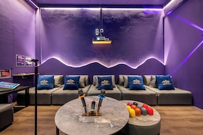 Quarto em tons de roxo, com área de estar, mesa e suporte para microfone, e um projetor fixado na parede.