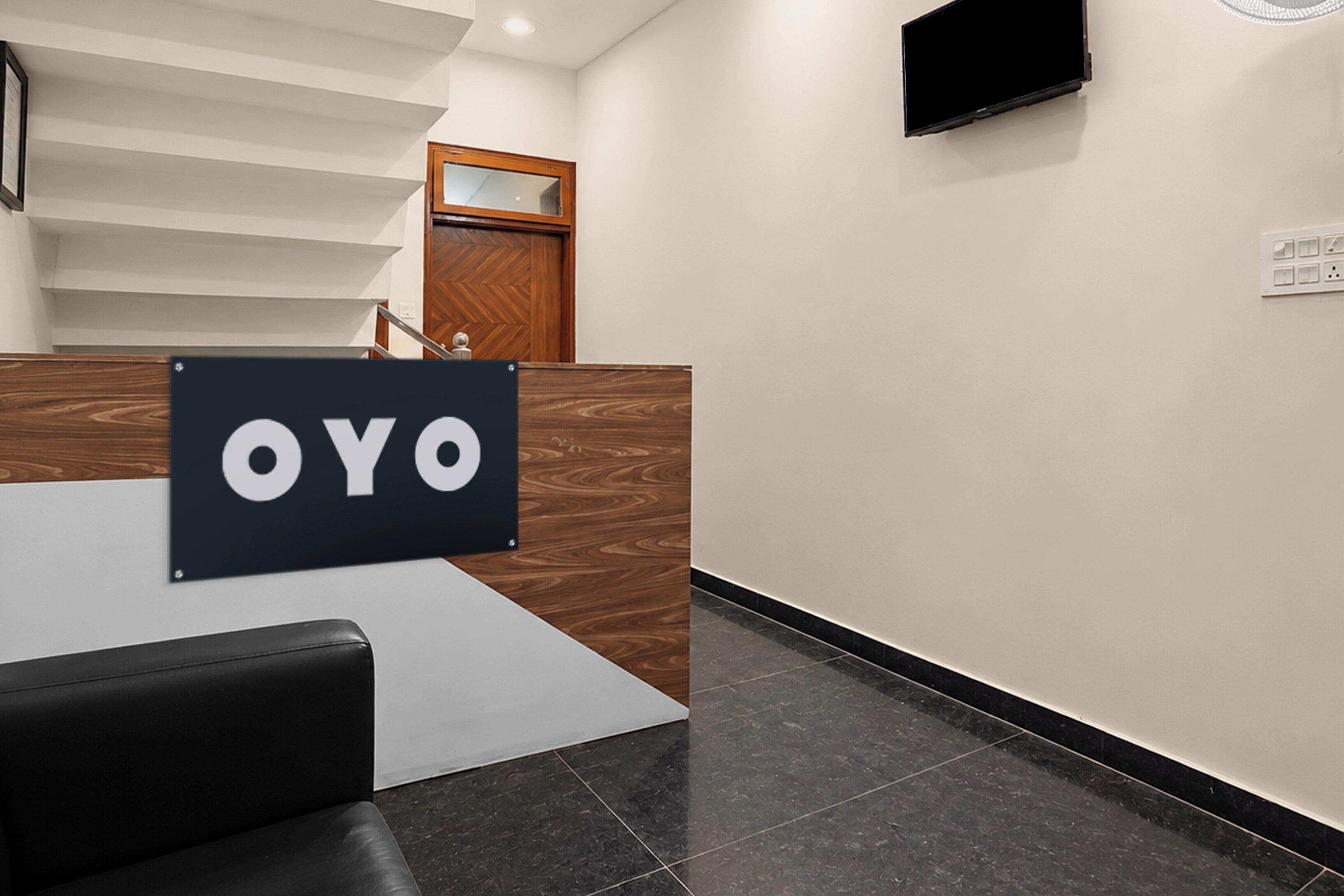 Uma receção com uma placa a indicar 'OYO' num hall moderno de hotel, com uma escadaria e uma televisão de ecrã plano na parede.