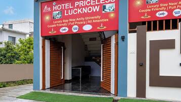 Entrada de hotel com uma placa vermelha anunciando 'Hotel Priceless Lucknow' e oferecendo acomodações de luxo, ideal para casais, privacidade total e todas as comodidades.