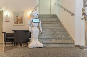 Um robô com uma tela exibindo 'Hotel 2.0 Kinder' em um saguão com escada e um quadro na parede.