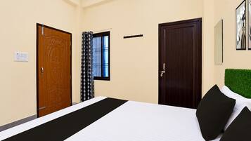 Quarto de hotel com cama, duas portas, janela com cortinas, interruptor de luz fixado na parede e uma pequena planta verde.