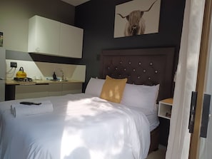 Quarto de hotel com cama, mesa de cabeceira, quadro na parede e cozinha equipada com chaleira e pia.