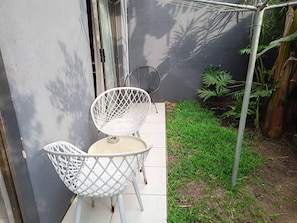 Uma área de pátio com uma mesa de metal branca e duas cadeiras, um pequeno jardim com grama e plantas, e uma parede cinzenta.