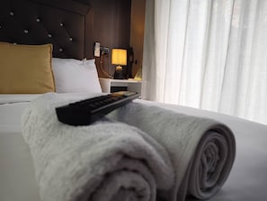 Cama bem arrumada com um controle remoto em cima, luminária de cabeceira e mesa de cabeceira com um relógio.