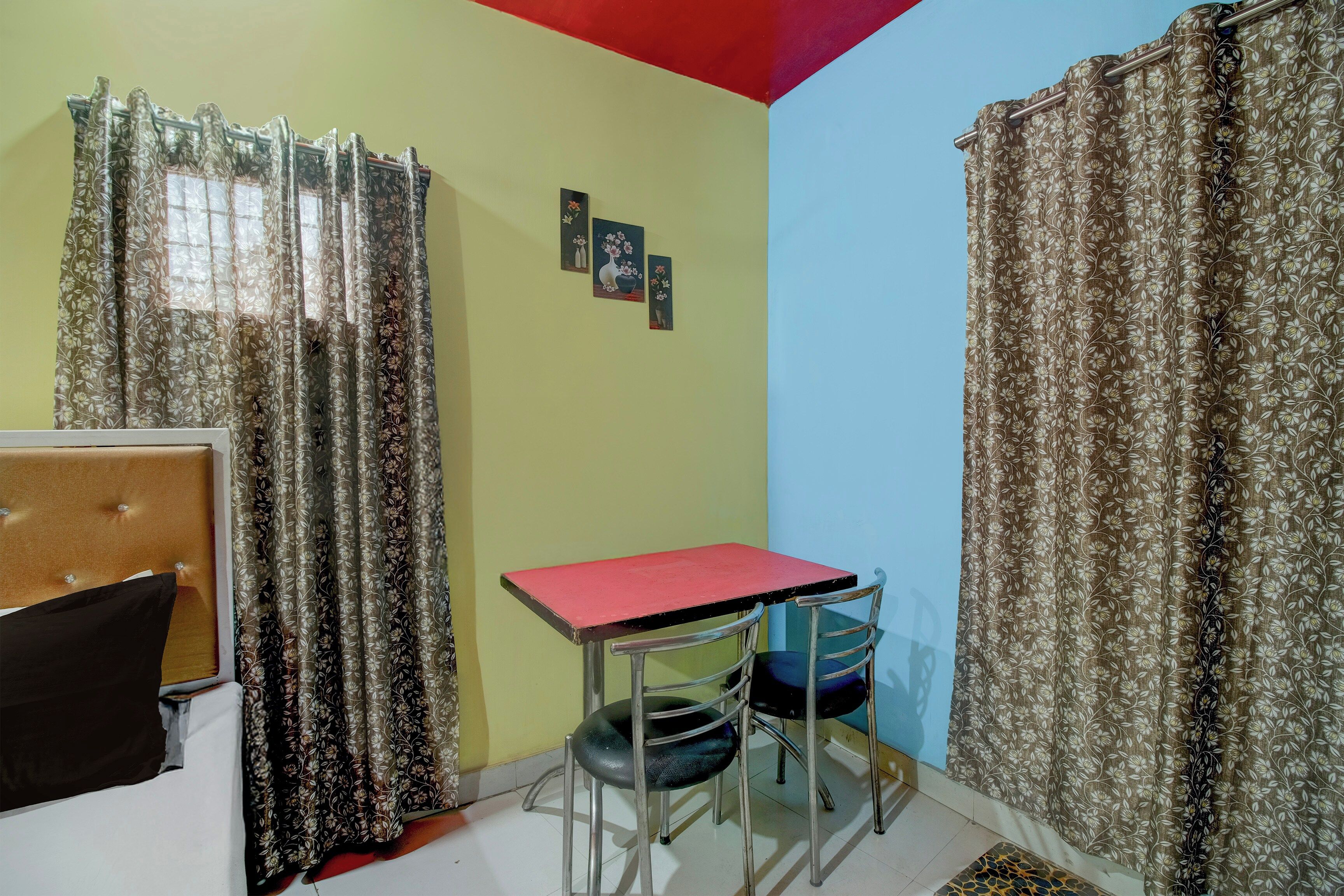 Quarto pequeno com cama, mesa e cadeira. Cortinas nas janelas.