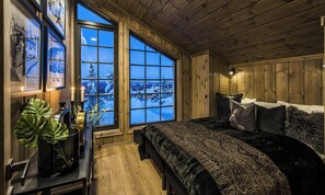Quarto com cama, mesa de cabeceira, janela com vista para uma paisagem nevada e quadros na parede.