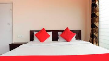 Quarto de hotel com uma cama, duas almofadas vermelhas, cabeceira de madeira, parede branca e janela com cortinas estampadas.