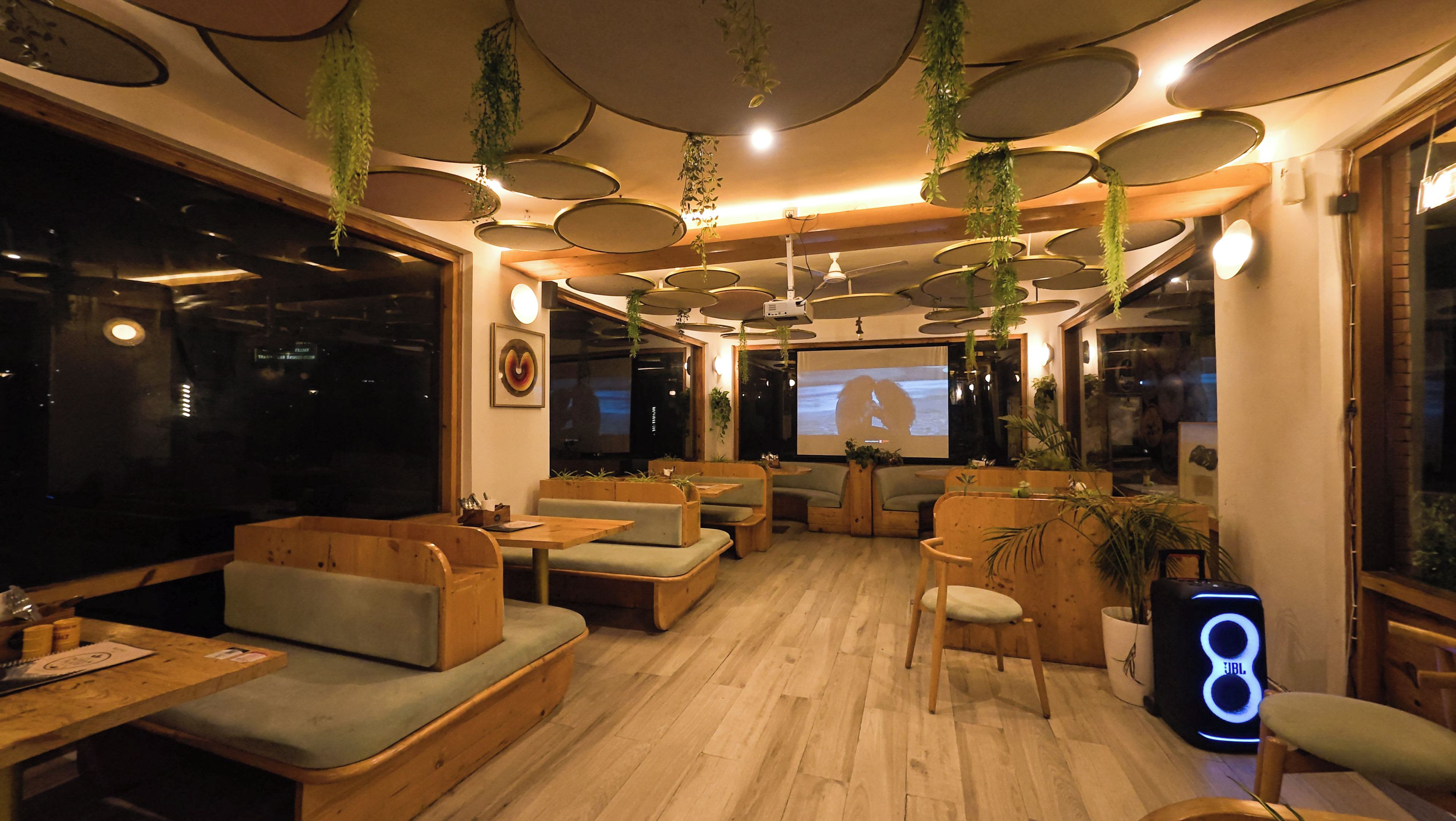 Interior acolhedor de restaurante, com mobiliário de madeira, plantas penduradas e uma tela de projeção.