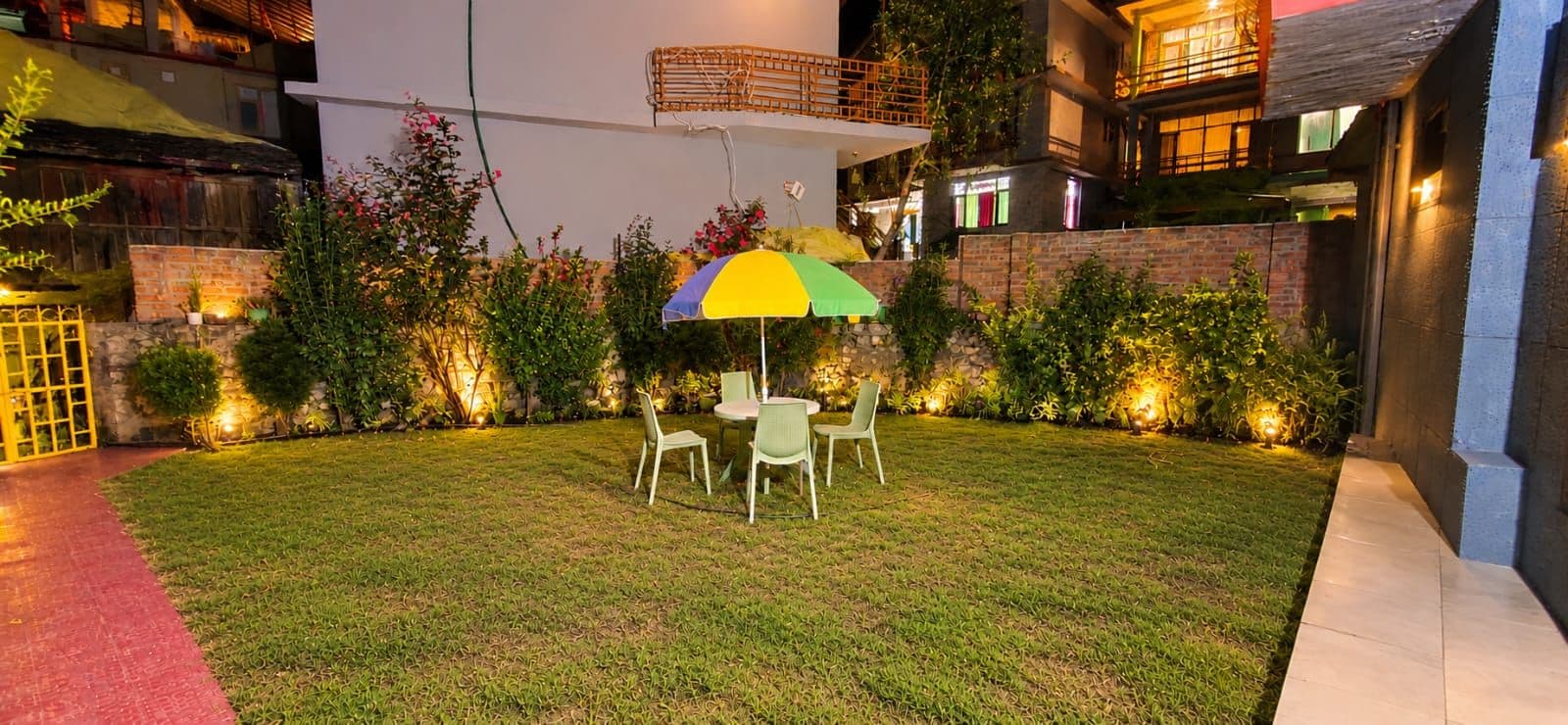 Área externa bem iluminada com gramado, mesa e cadeiras, e um guarda-sol colorido.
