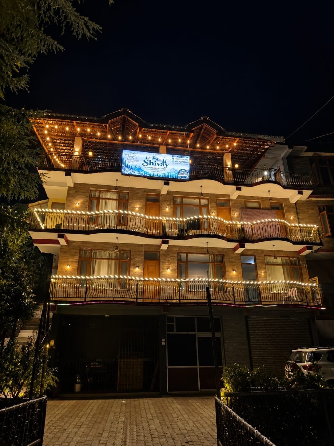 Hotel com vários andares, varandas decoradas com luzes de Natal e área de entrada pavimentada, com um letreiro digital em destaque.