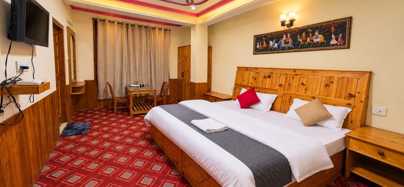 Quarto de hotel com uma cama grande, cabeceira de madeira, televisão de tela plana na parede, escrivaninha com cadeira e um quadro sobre a cama.