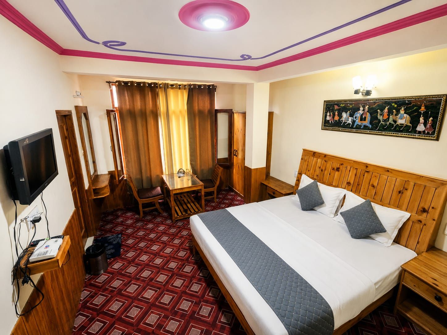 Quarto de hotel com cama, escrivaninha, cadeira, televisão e um quadro na parede.