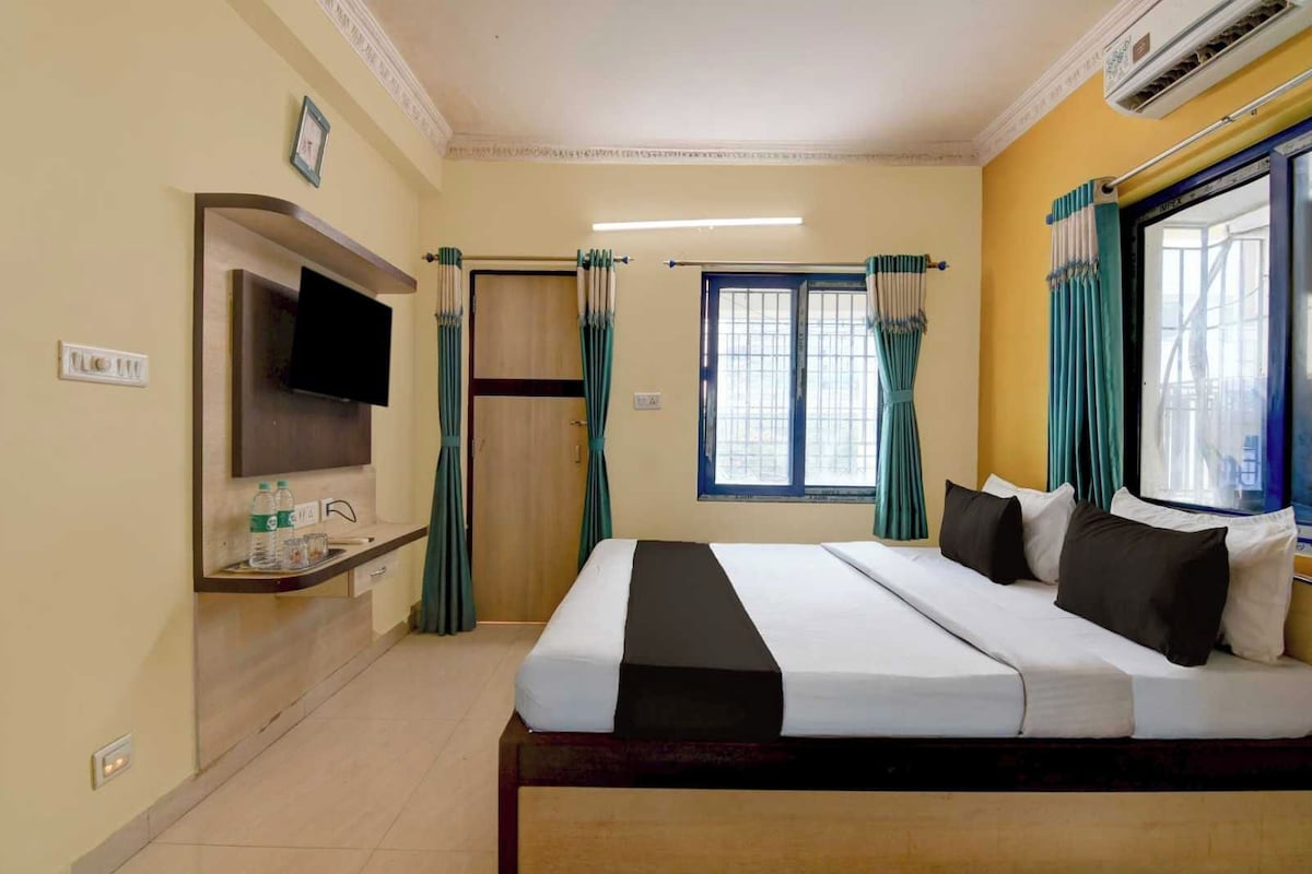 Quarto de hotel com cama, televisão, janela com cortinas e uma pequena mesa com garrafas de água.