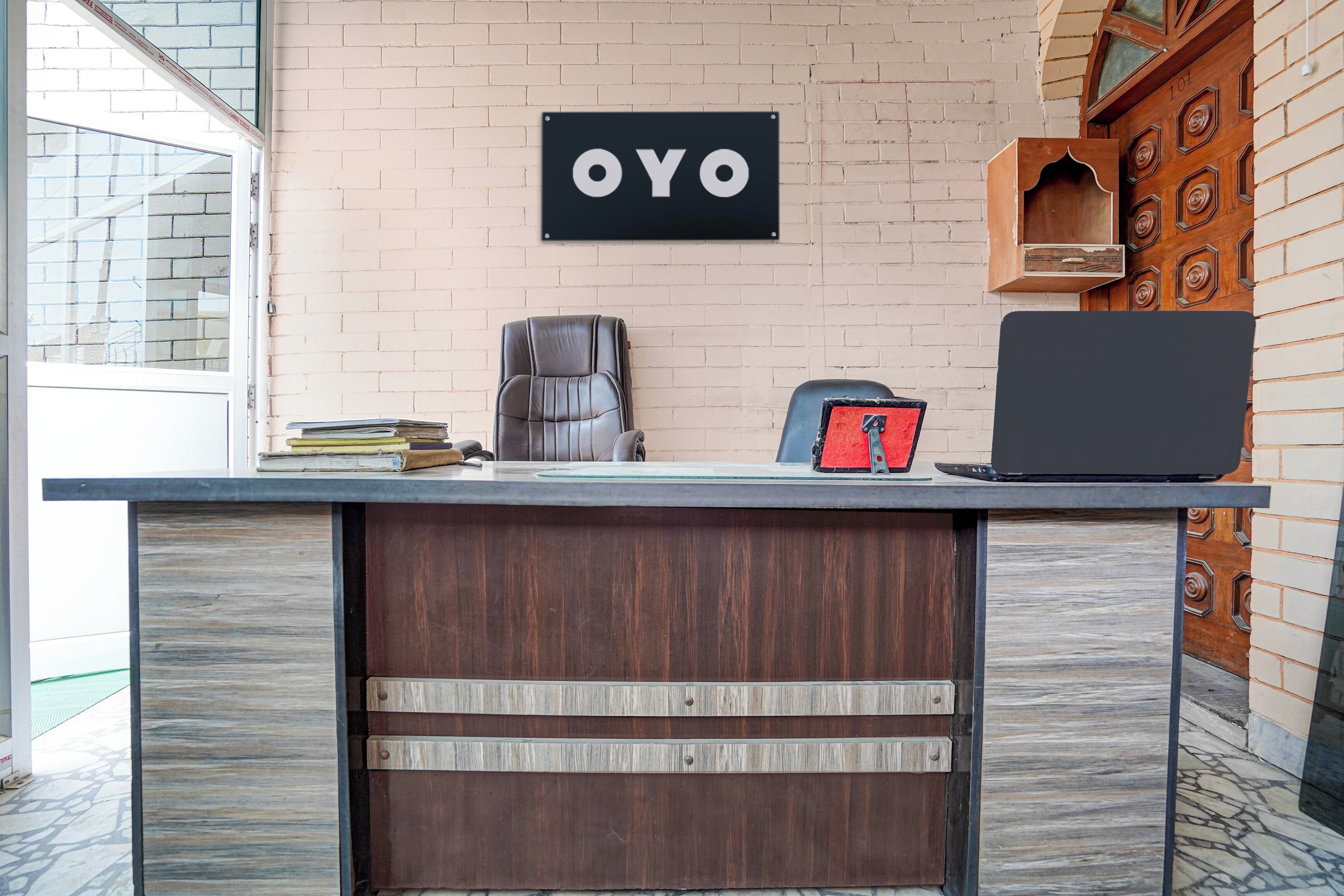 Bancada de recepção com placa indicando 'OYO', uma poltrona de couro, um laptop e um objeto vermelho no balcão.