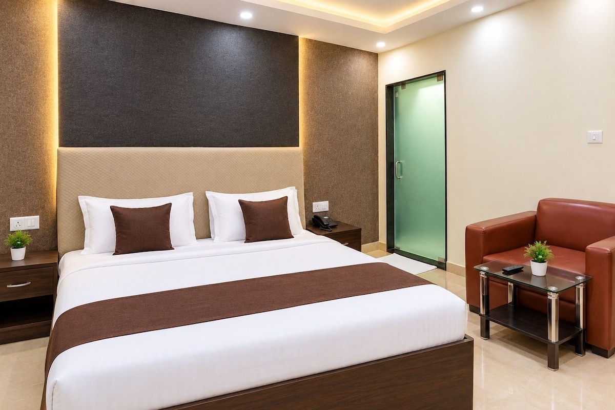 Quarto de hotel com uma cama grande, uma escrivaninha com cadeira, uma porta verde e uma pequena mesa com uma planta.