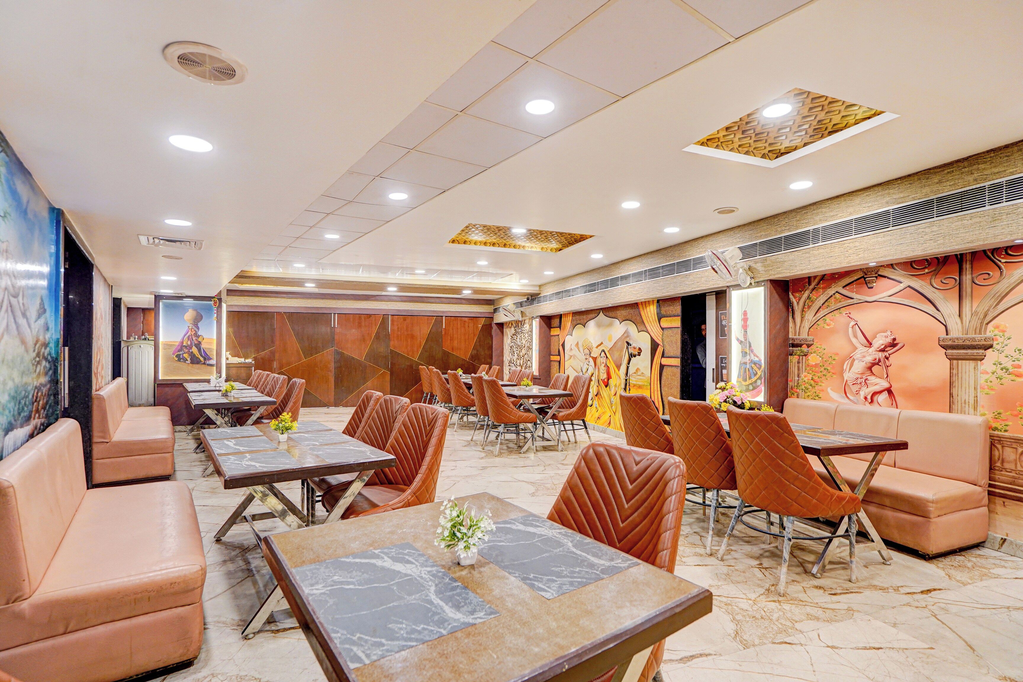 Restaurante com várias mesas e cadeiras, teto com iluminação embutida e murais decorativos nas paredes.