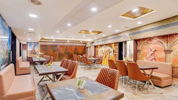 Restaurante com várias mesas e cadeiras, teto com iluminação embutida e murais decorativos nas paredes.